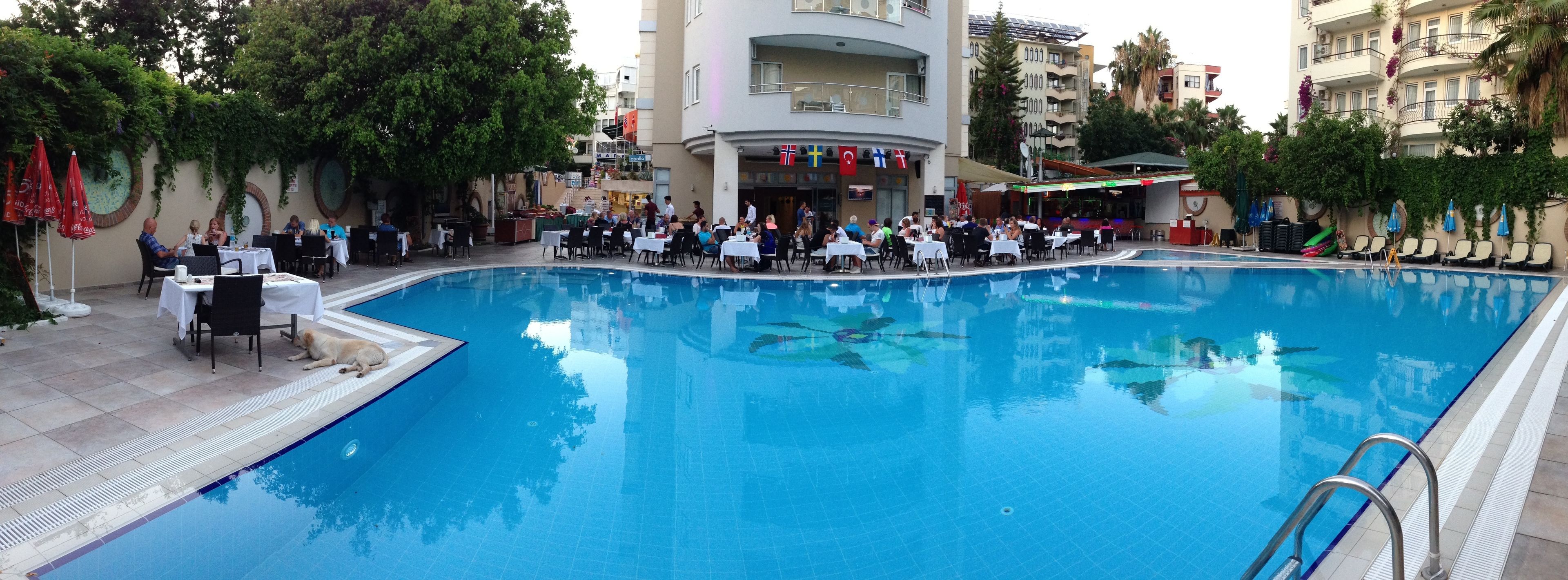 Piscina com mesas e cadeiras ao redor, um edifício com varandas e bandeiras.