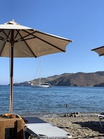 Uma praia com guarda-sóis, um trecho de costa rochosa e um veleiro ao fundo.