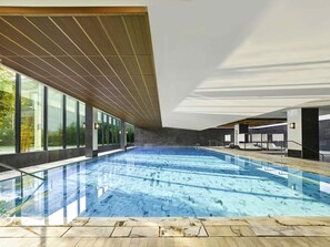 Uma piscina interior moderna com paredes de vidro transparente, chão revestido de azulejos e teto de madeira.