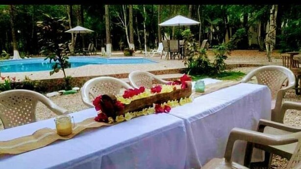 Uma área de refeições à beira da piscina, com uma mesa posta para dois, cadeiras ao redor e uma piscina ao fundo.