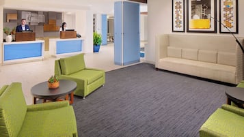 Um hall de hotel com balcão de recepção, poltronas verdes, um sofá bege e piso coberto por carpete.