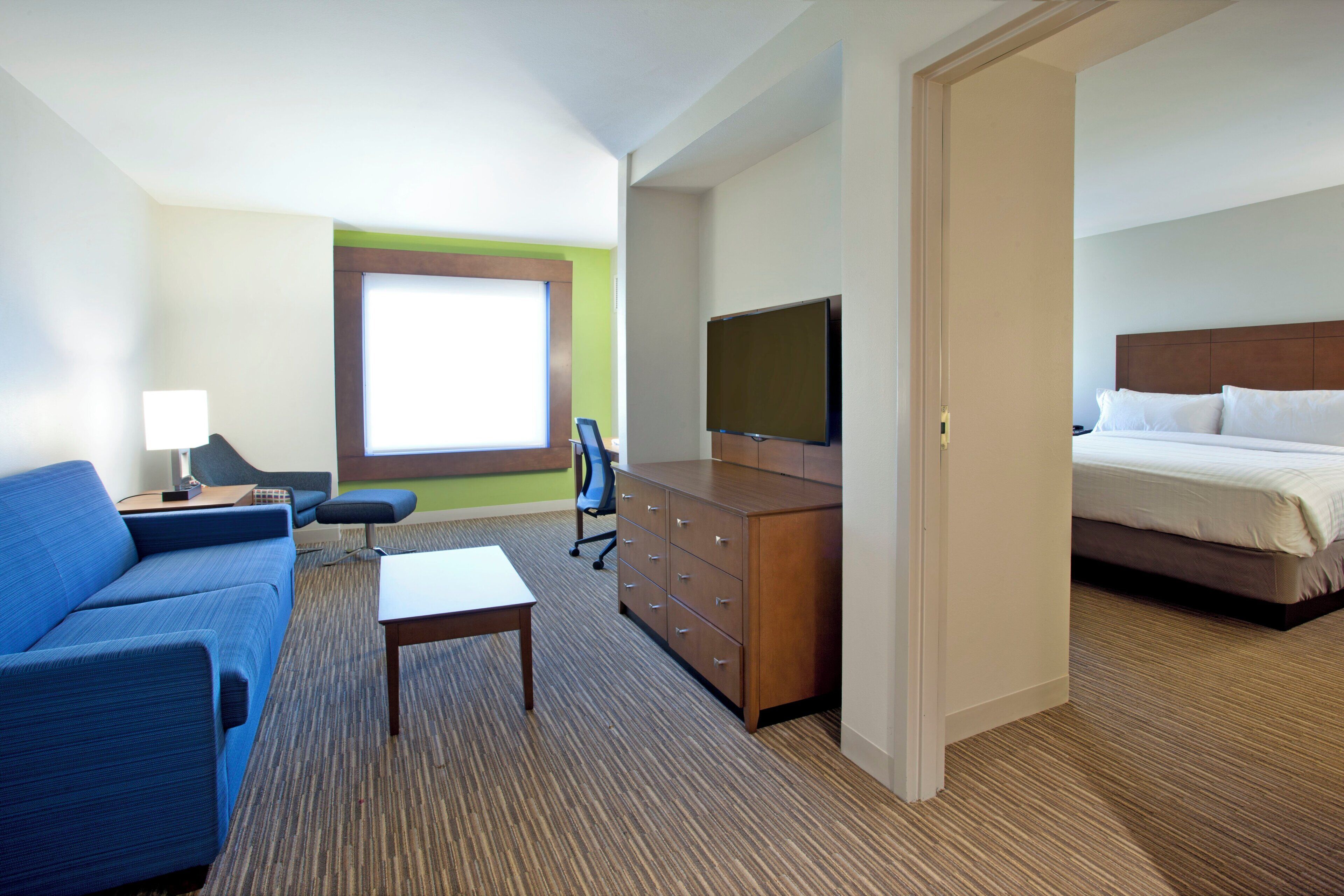Quarto de hotel com cama, escrivaninha, cadeira, televisão e sofá.