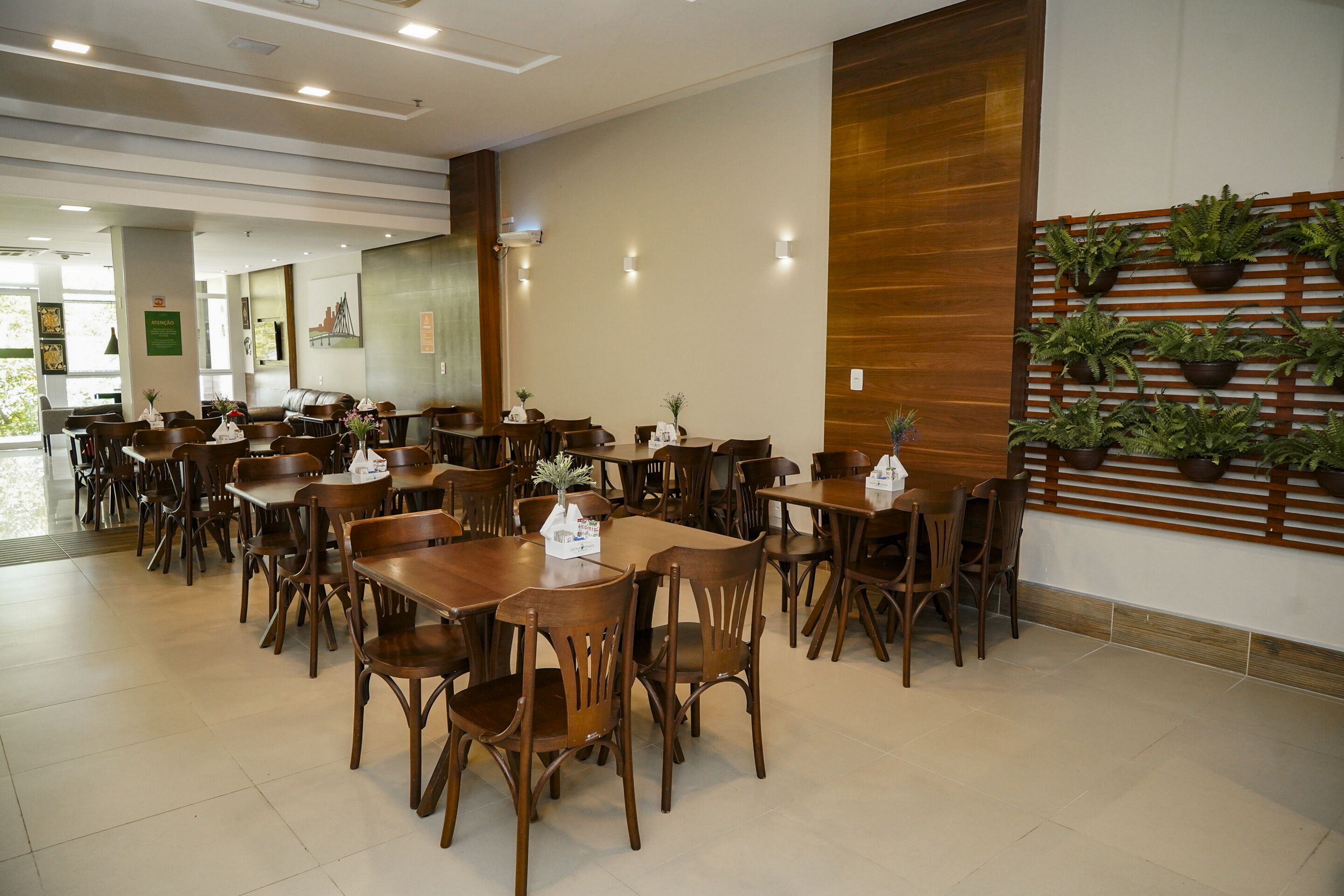 Uma ampla sala de jantar com mesas e cadeiras de madeira, um painel de madeira decorado com plantas em vasos, e vista para o exterior por grandes janelas.