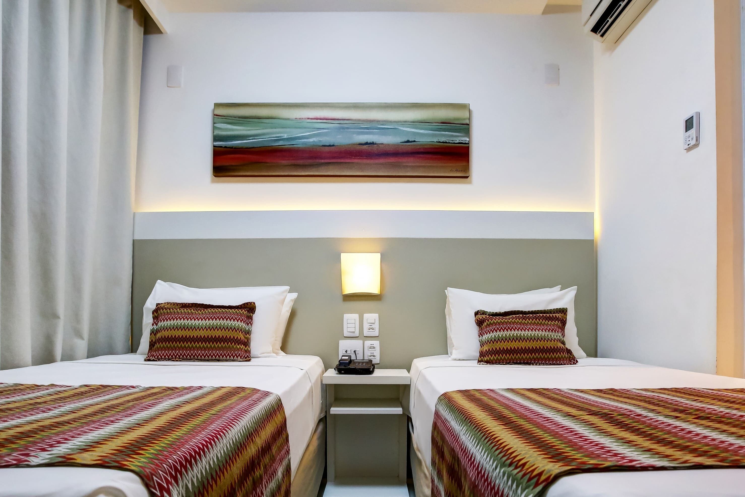 Quarto de hotel com duas camas, uma mesa de cabeceira, um quadro na parede e uma luminária no teto.