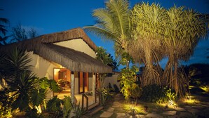 Uma casa com telhado de palha, rodeada por plantas tropicais e um caminho de pedra.