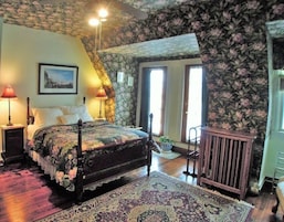 Quarto com cama de dossel, papel de parede floral, duas luminárias de cabeceira, uma janela grande e piso de madeira.