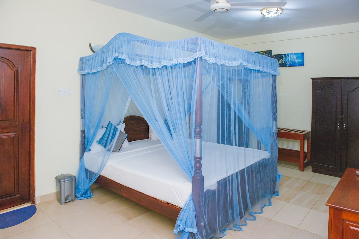 Quarto com cama sob um dossel azul, um armário de madeira e uma pequena lixeira.