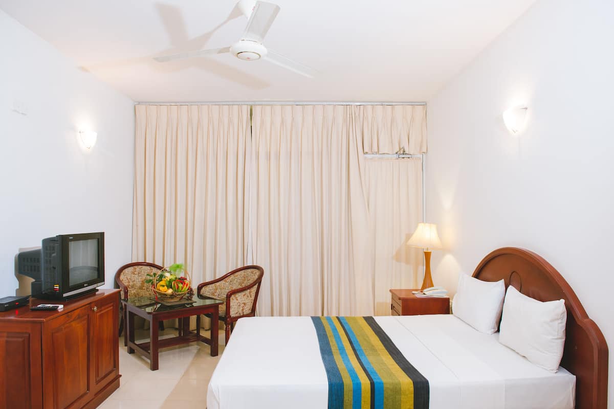 Quarto de hotel com cama, televisão, escrivaninha com abajur, cadeira e ventoinha no teto.