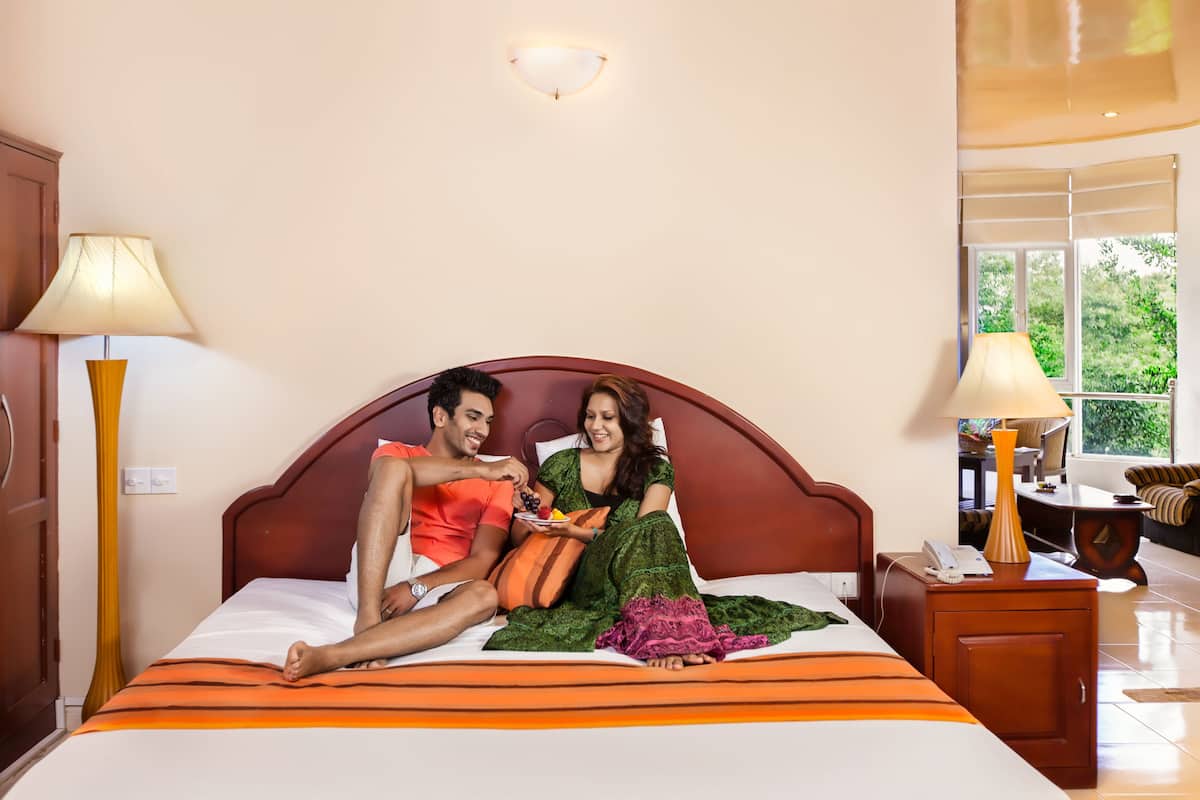 Quarto com cama, mesa de cabeceira, luminária e janela com vista para área verde.