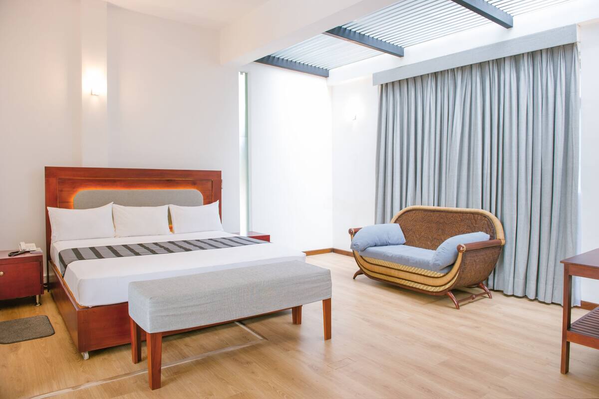 Quarto de hotel com cama de madeira, um banco, uma cadeira de vime e uma janela grande com cortinas.
