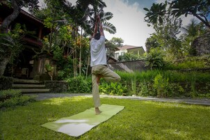 Uma pessoa fazendo uma postura de yoga em um tapete, em um jardim exuberante, com uma casa ao fundo.