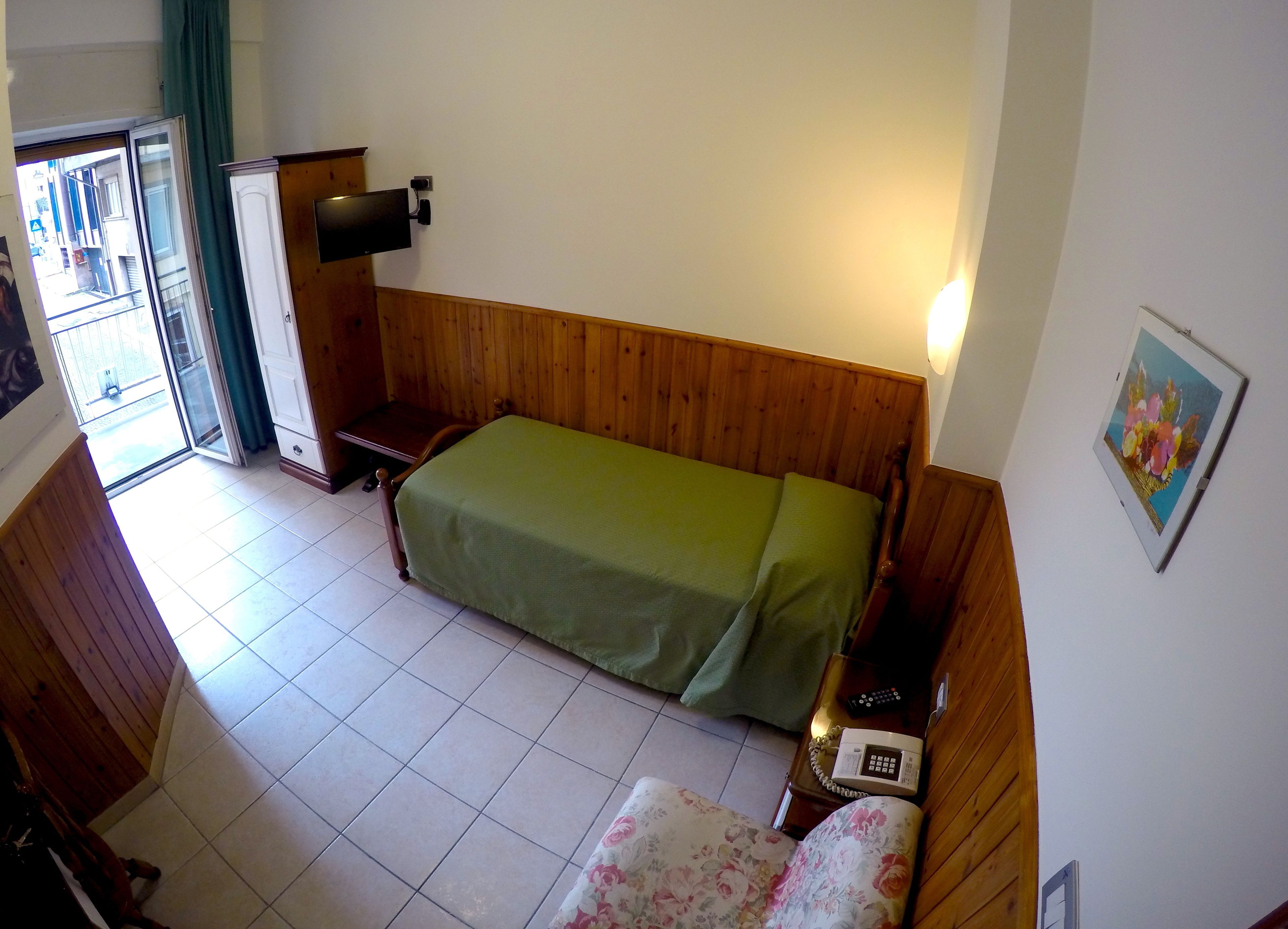 Quarto pequeno com uma cama de solteiro, um guarda-roupa de madeira, uma mesinha com telefone e uma luminária na parede.