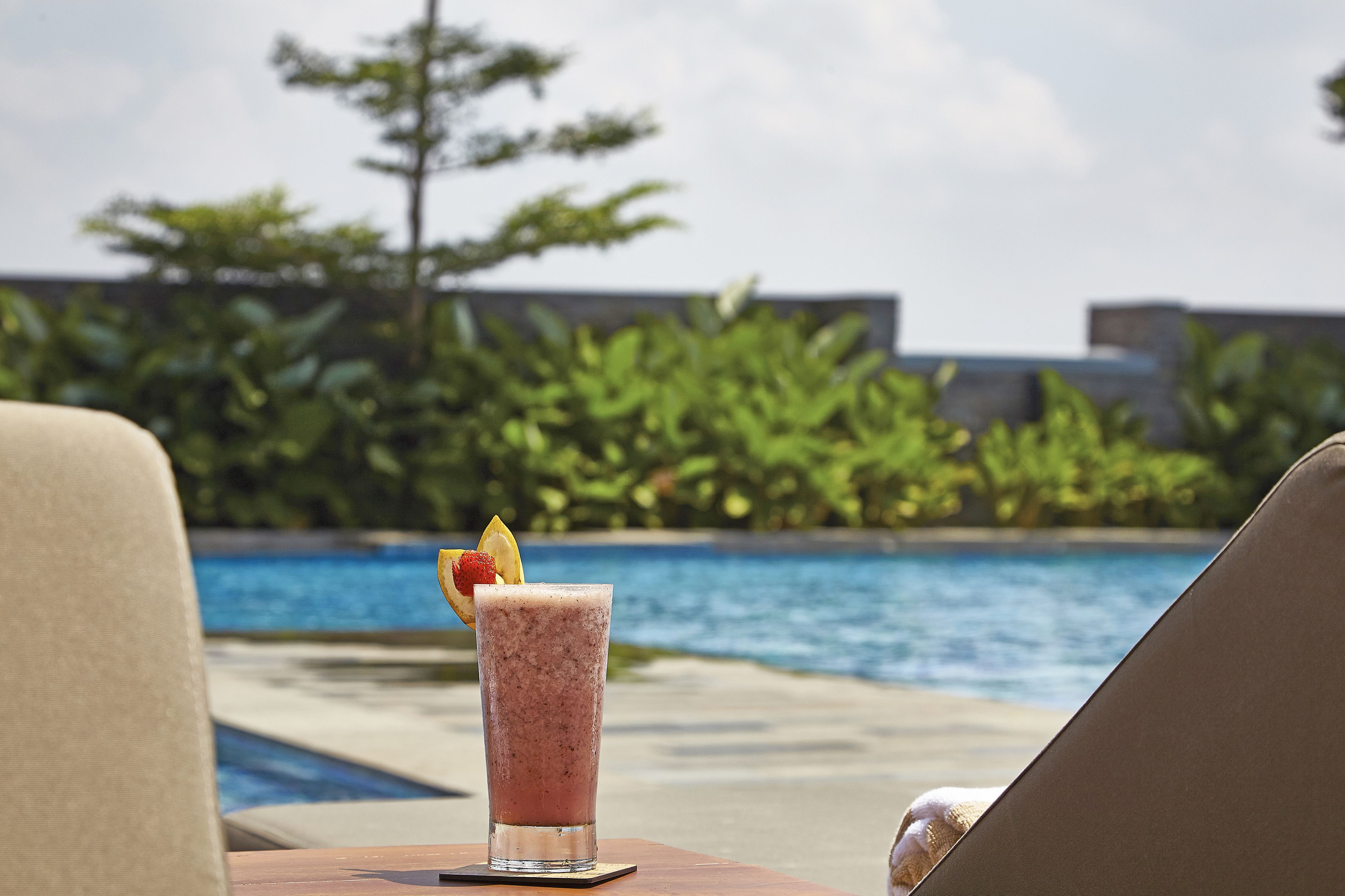 Um copo de smoothie com decoração de fruta sobre uma mesa perto de uma espreguiçadeira à beira da piscina.