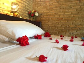 Cama com lençóis brancos e pétalas de rosa vermelhas espalhadas, com uma parede de tijolos e um aparelho de luz decorativo acima.