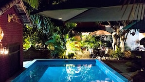 Piscina com parede de tijolos de um lado e área de varanda coberta com cadeiras e guarda-sóis.