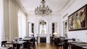 Uma sala de jantar formal com lustres, paredes brancas e lareira. Mesas postas com copos e talheres.