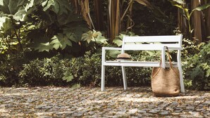 Um banco branco com uma bolsa trançada em um jardim com grandes folhas de bananeira e um caminho de pedras.