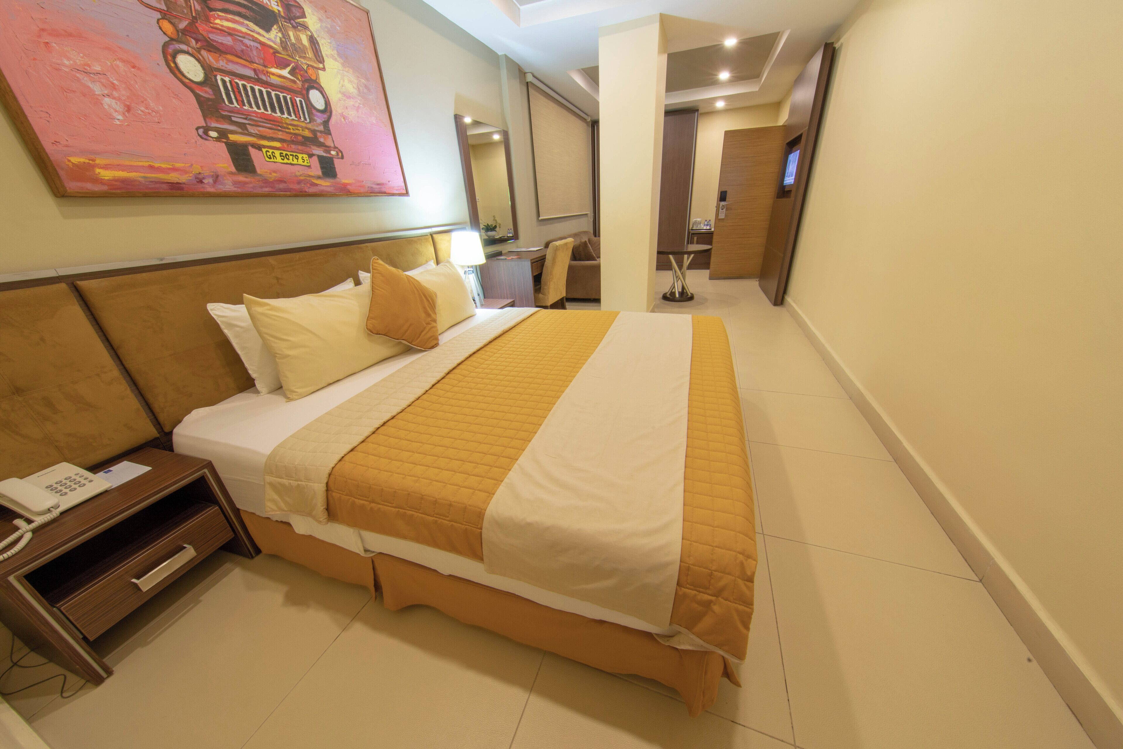 Quarto de hotel com uma cama grande, mesa de cabeceira com telefone, escrivaninha com cadeira e um quadro na parede.