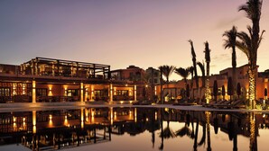 Resort com piscina que reflete os edifícios e palmeiras, com área de estar ao ar livre e espaço coberto.