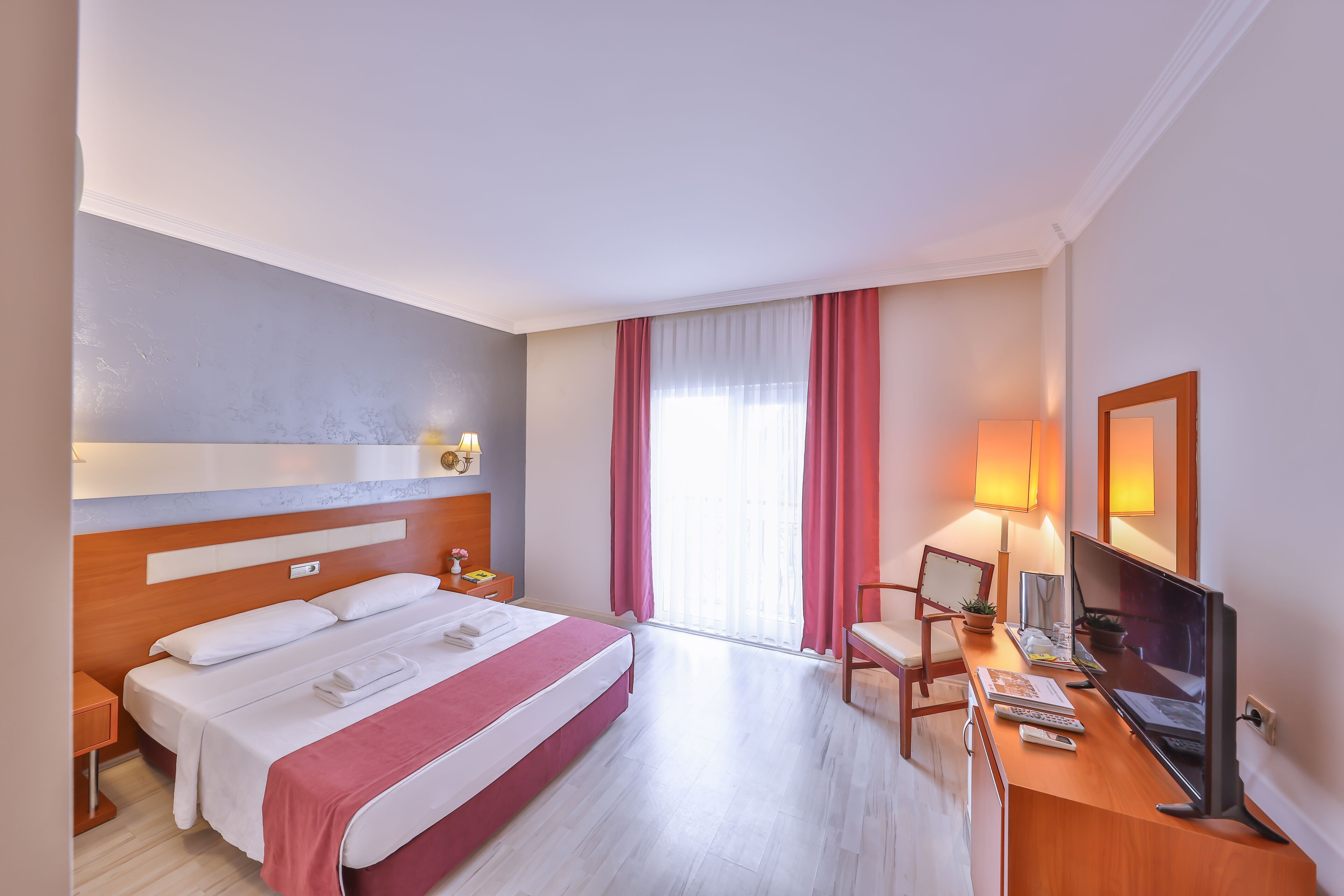 Quarto de hotel com cama, mesa de cabeceira, abajur, televisão e escrivaninha com cadeira.