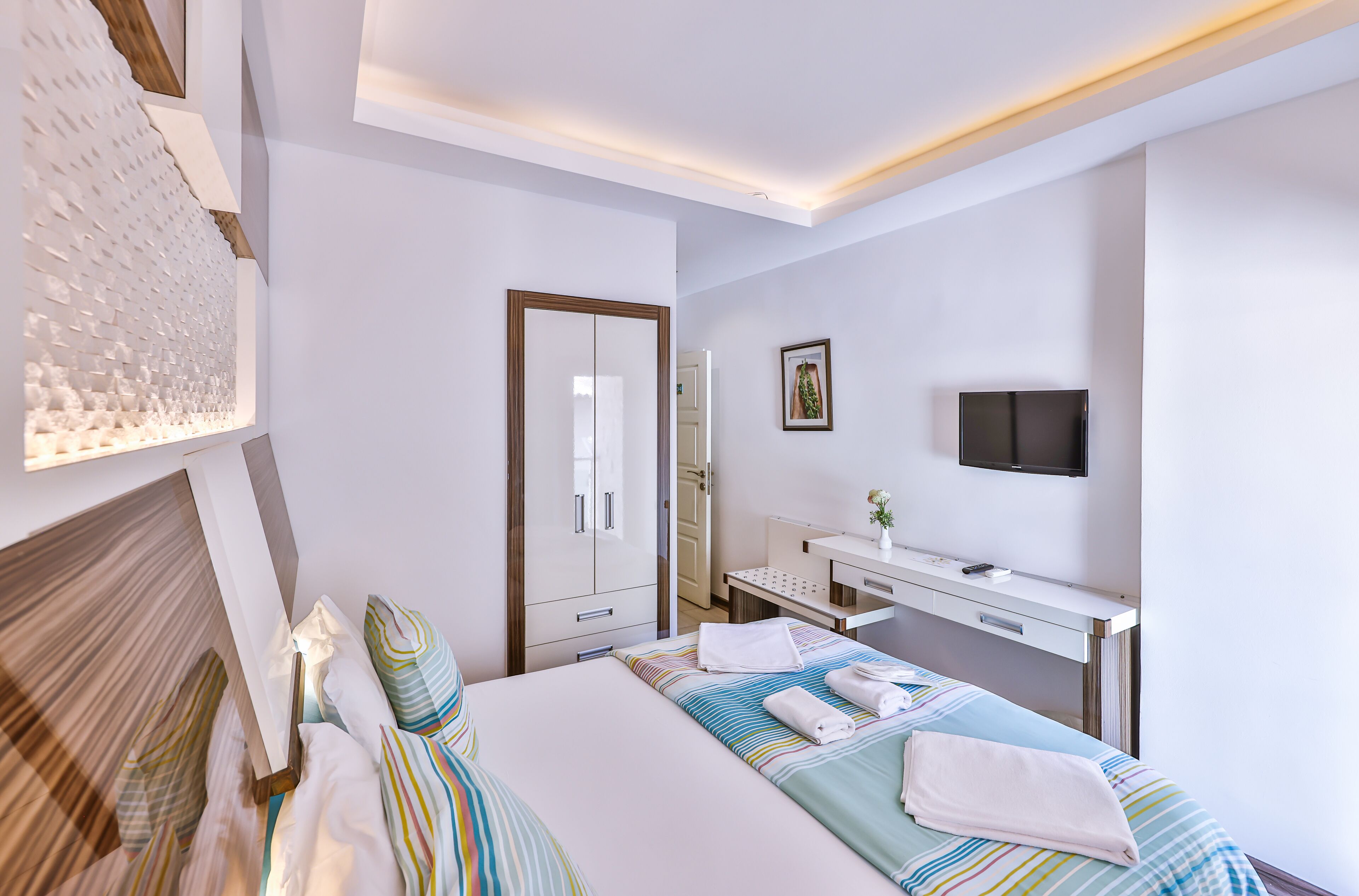 Quarto de hotel com cama, mesa de cabeceira, escrivaninha e televisão.