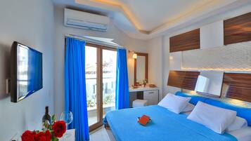 Quarto de hotel com cama, televisão, escrivaninha, banheiro e varanda com cortinas azuis.