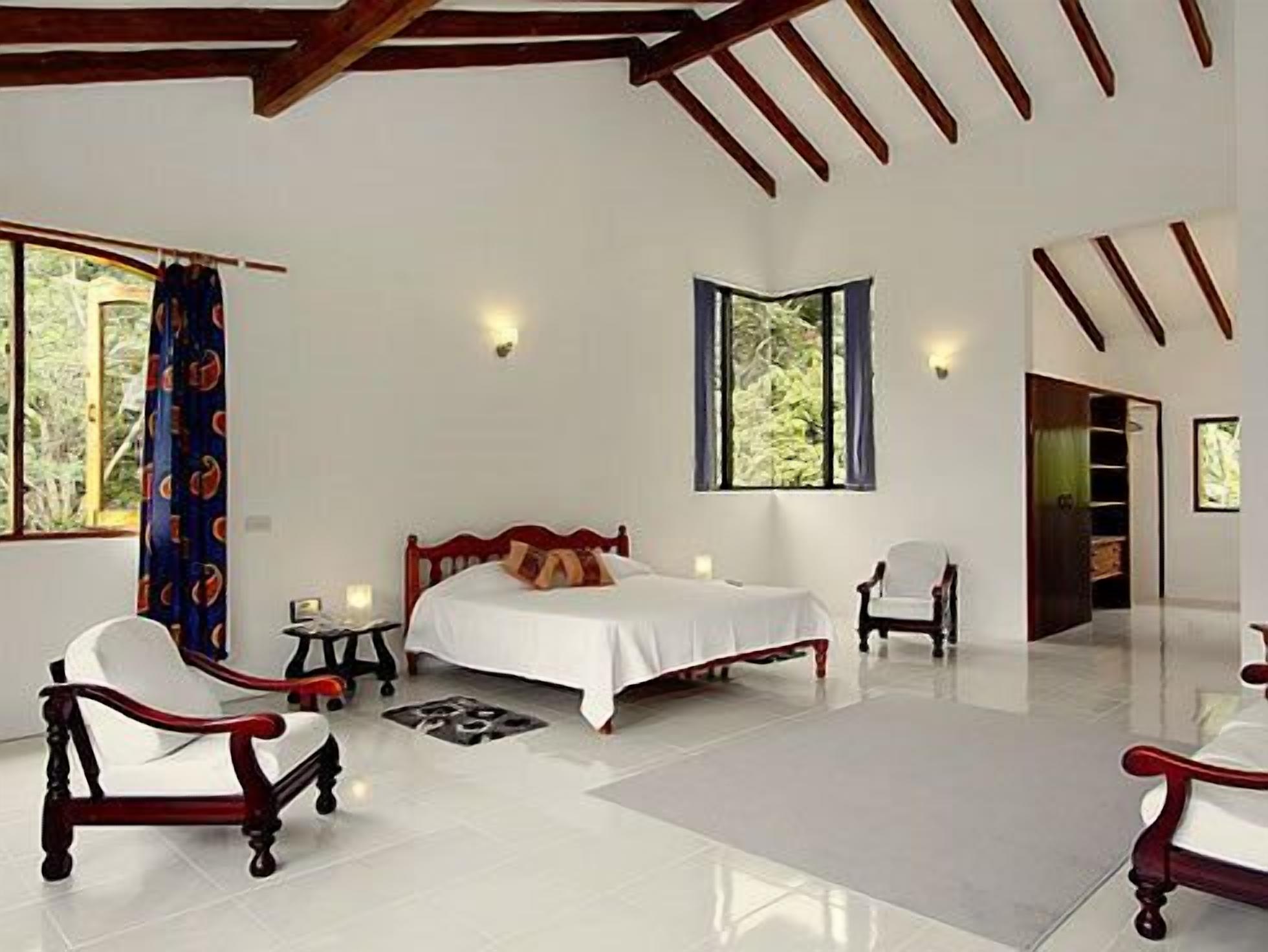 Quarto com uma cama, duas cadeiras, mesa de cabeceira e janela com cortinas.