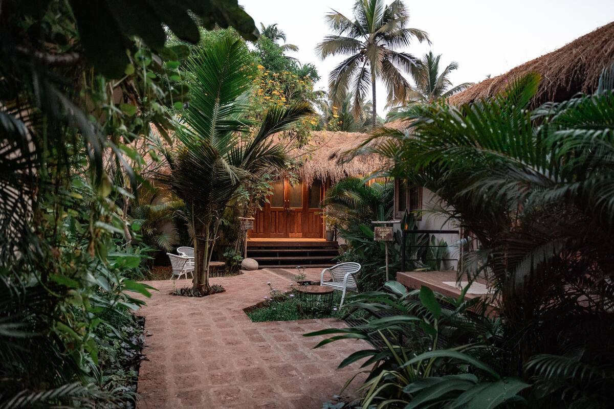 Um caminho em um jardim tropical leva a uma porta de madeira com telhado de palha, cercado por vegetação exuberante e palmeiras.