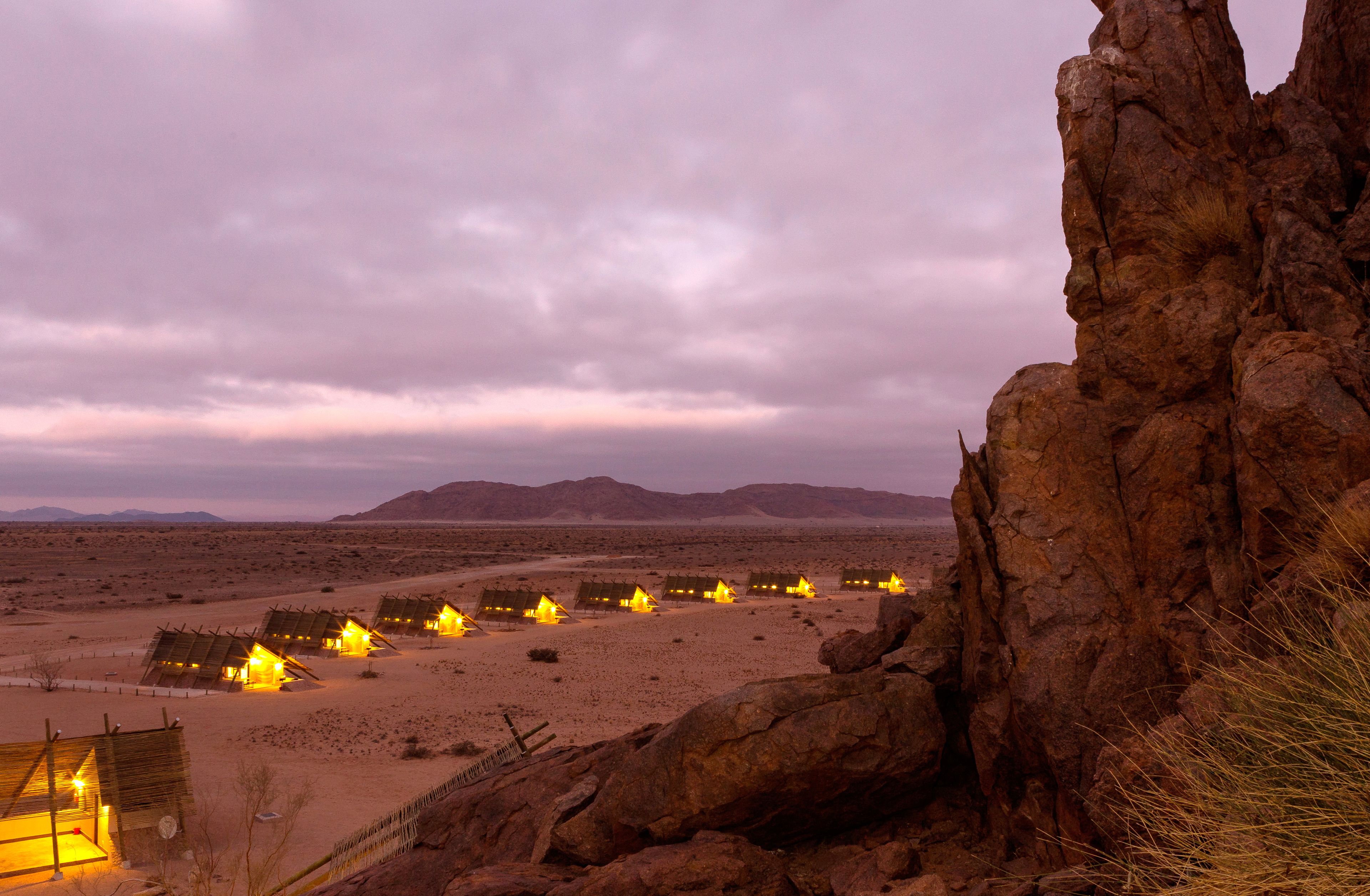 Paisagem desértica com várias estruturas pequenas iluminadas, provavelmente alojamentos, dispostas em linha.