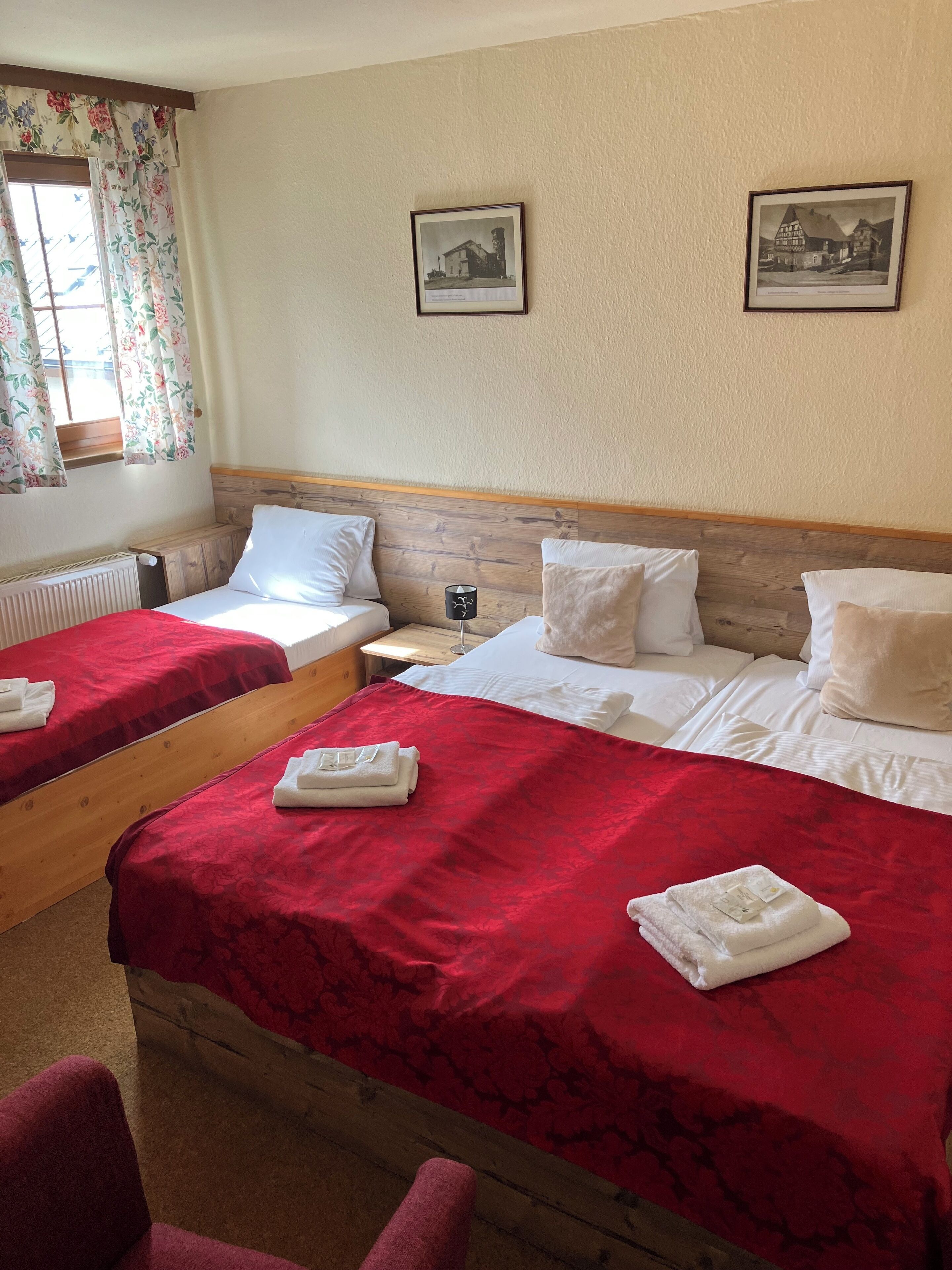 Quarto de hotel com duas camas, cada uma com roupa de cama vermelha e almofadas brancas, uma cabeceira de madeira, uma mesa de cabeceira com abajur e duas fotos emolduradas na parede.