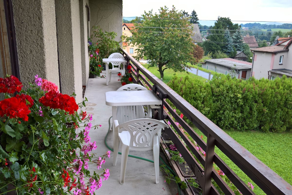 Varanda com mesa e cadeira brancas, cercada por flores vermelhas e cor-de-rosa, e vista para uma paisagem verde e edifícios ao fundo.
