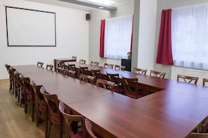 Uma sala de conferências com uma longa mesa de madeira, várias cadeiras, uma tela de projetor e janelas com cortinas vermelhas.