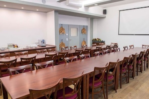 Uma sala de conferências com longas mesas e cadeiras de madeira, um espaço para café e uma tela de projetor na parede.