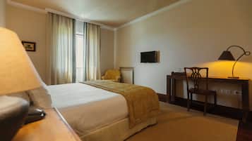 Quarto de hotel com cama, escrivaninha, cadeira, abajur e televisão na parede.