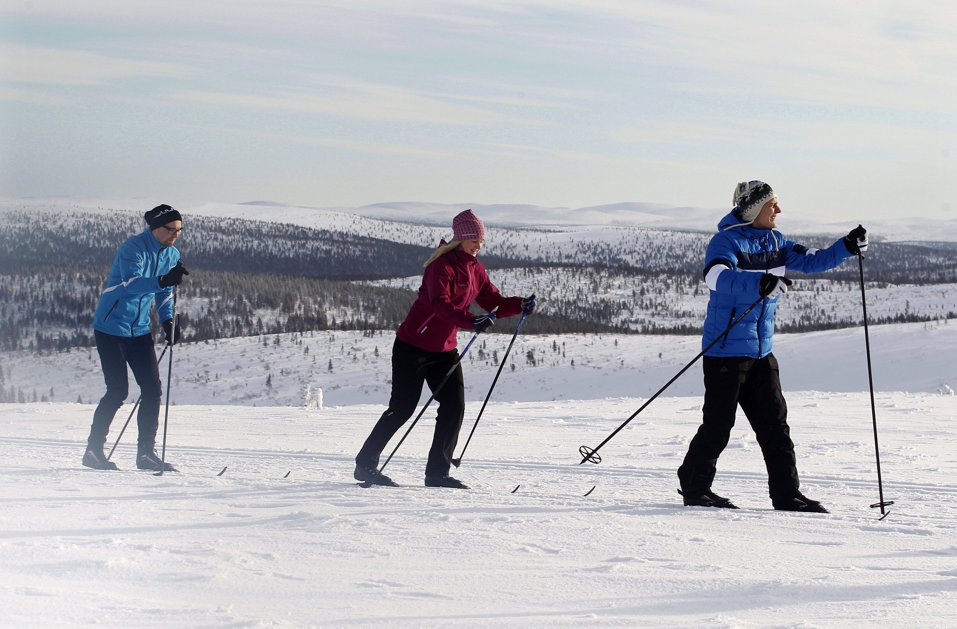 Três pessoas praticam esqui cross-country em uma paisagem nevada, com árvores e colinas ao fundo.