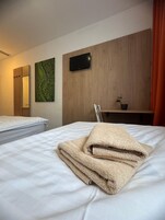 Quarto de hotel com cama, televisão fixada na parede e uma pequena mesa com uma planta.