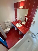 Um banheiro com banheira branca, duas pias, vaso sanitário e parede com azulejos vermelhos.