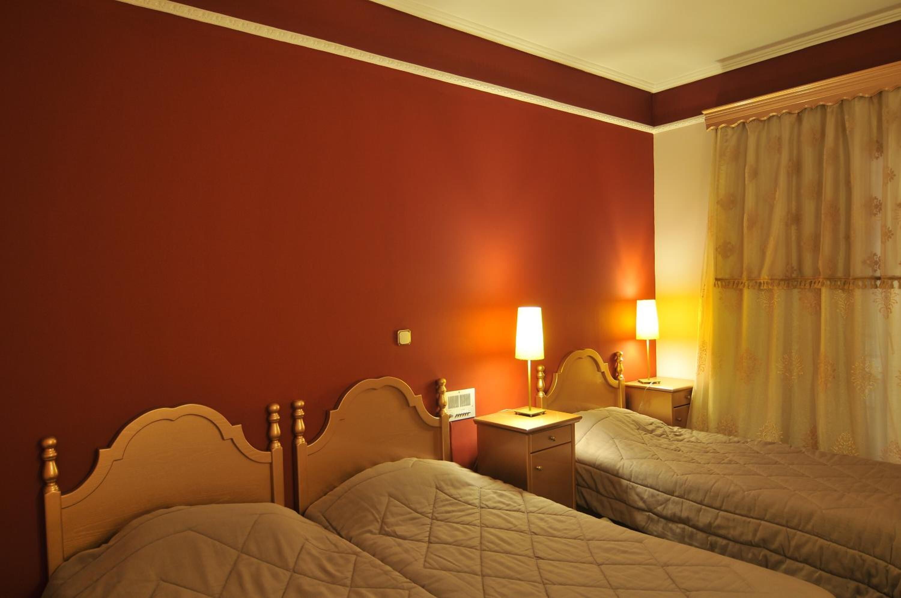 Quarto de hotel com duas camas, uma mesa de cabeceira, uma luminária e uma janela com cortinas.