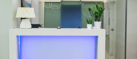 Um balcão de recepção com um painel azul iluminado, uma luminária branca e um vaso com planta.