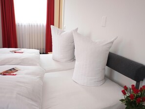 Quarto de hotel com cama, almofadas, mesa de cabeceira e janela com cortinas.