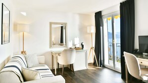 Quarto de hotel com sofá, escrivaninha, cadeira, espelho, abajur, televisão e varanda com vista.