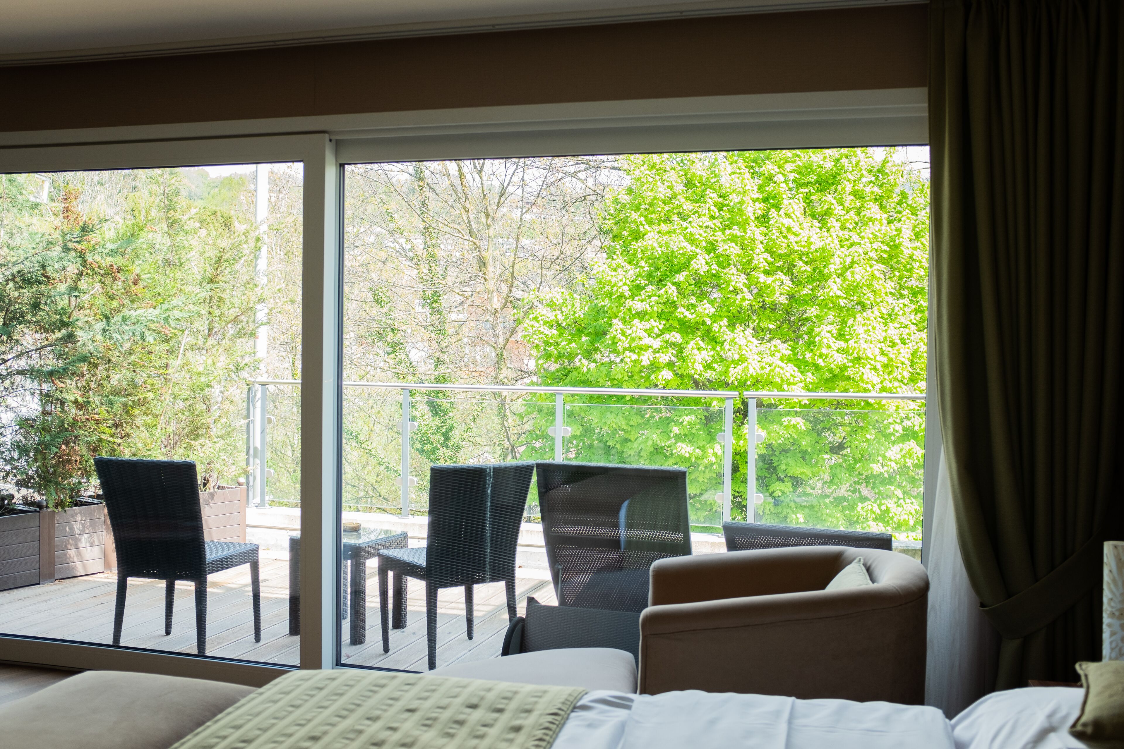 Quarto de hotel com uma cama, varanda com cadeiras de vime e uma mesa, e vista para o verde lá fora.