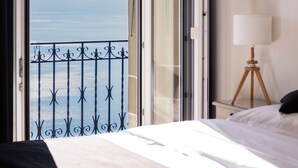 Quarto de hotel com cama, mesa de cabeceira, abajur e vista para o mar através de varanda com grade de metal.
