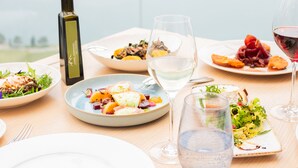 Uma mesa posta com vários pratos, copos de vinho e uma garrafa de vinho, com vista para uma paisagem montanhosa.