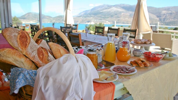 Mesa de café da manhã com pão, frutas e bebidas, com vista para uma paisagem montanhosa.