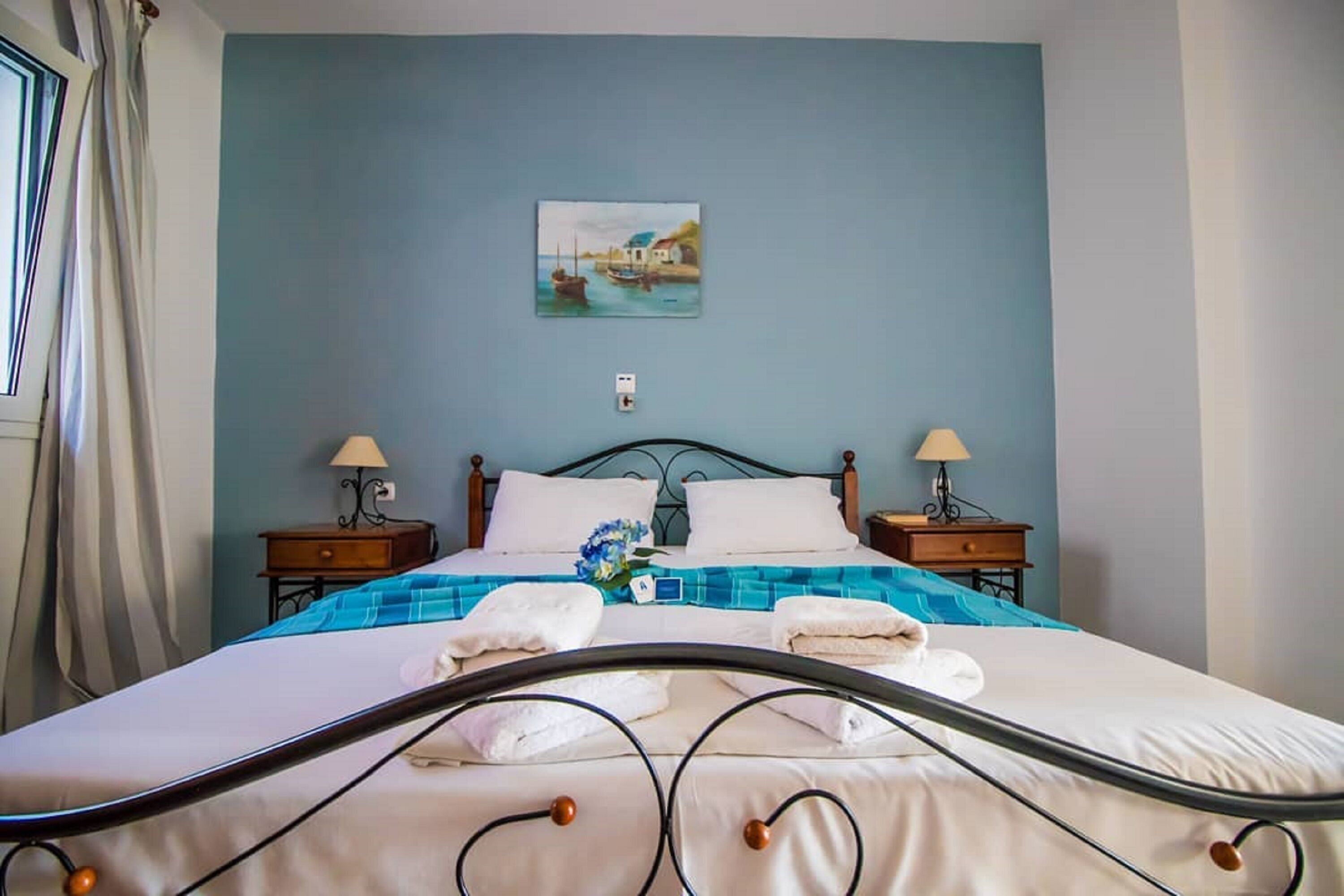 Cama bem arrumada com lençóis brancos e roupa de cama azul quadriculada, duas mesas de cabeceira com abajures, um quadro com cena costeira na parede e uma janela com cortinas.