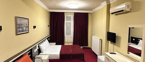 Quarto de hotel com cama, escrivaninha, cadeira, mesa pequena, espelho e ar condicionado.