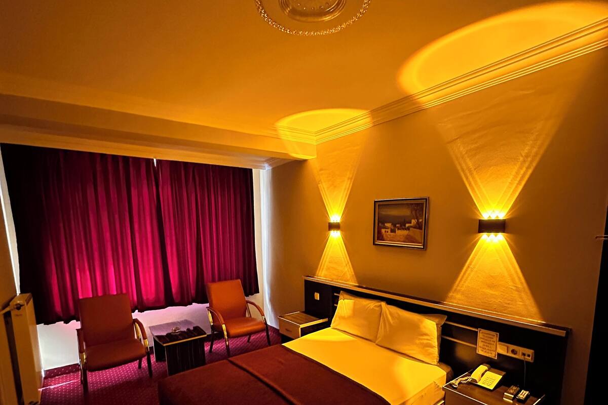 Quarto de hotel com uma cama grande, duas poltronas, uma escrivaninha e uma mesa de cabeceira com telefone.