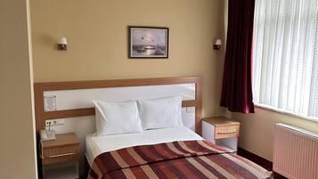 Quarto de hotel com cama, mesa de cabeceira, abajur, quadro e janela com cortinas.