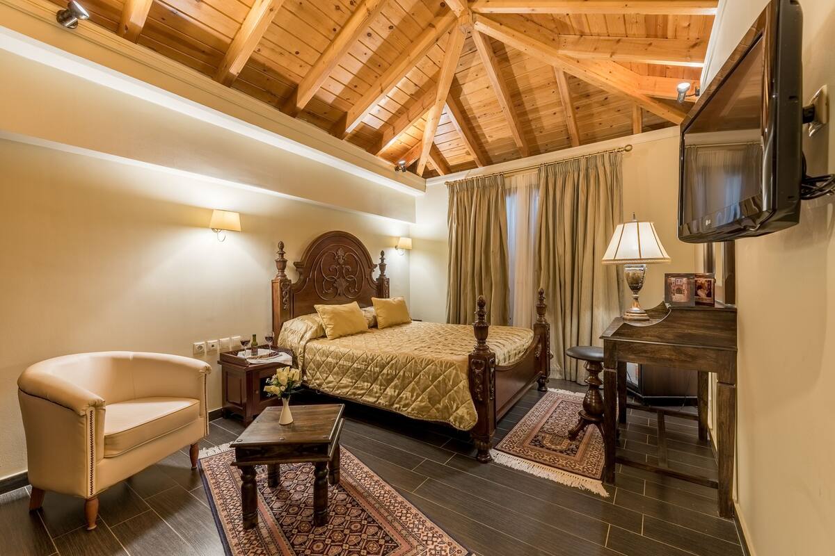 Quarto com teto de madeira, uma cama, um sofá, uma escrivaninha e uma televisão.